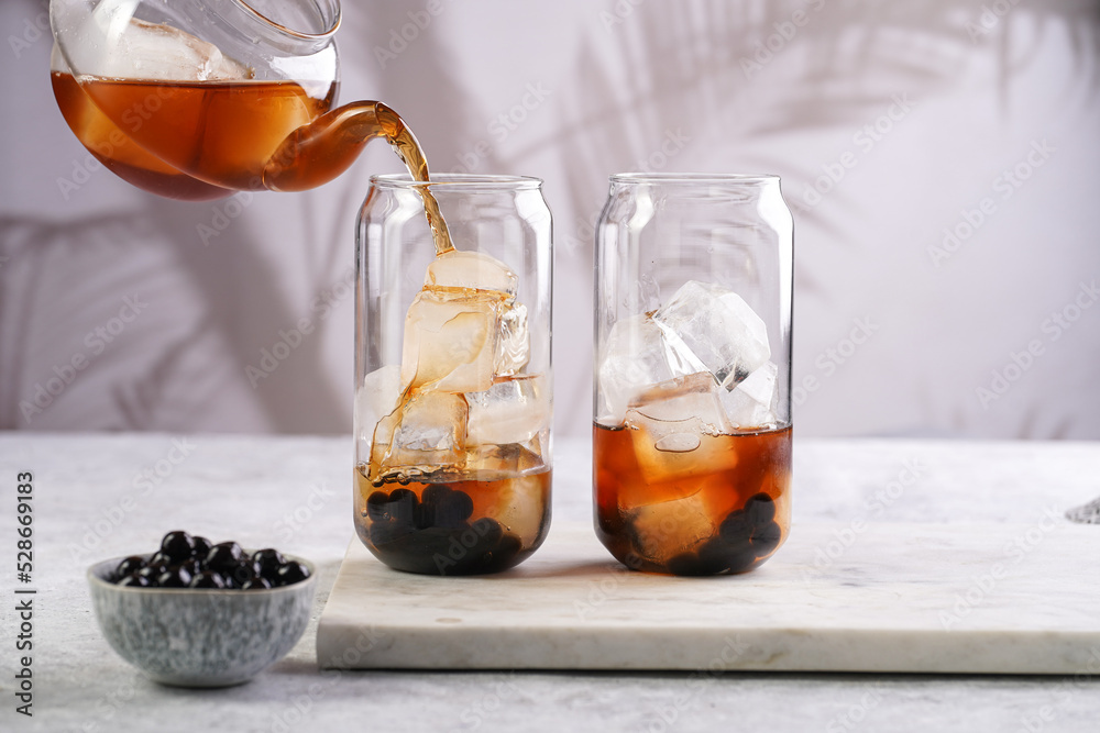 Female hand pouring black tea into transparent glass with ice cubes and ...