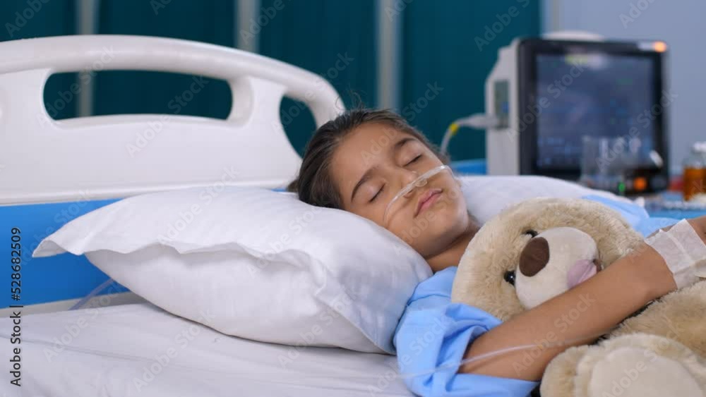 A young Indian girl sleeping in the hospital bed while hugging a teddy ...