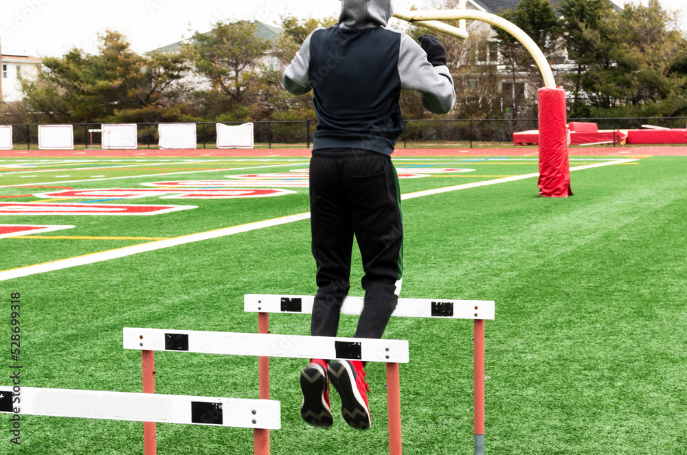 Athlete in fulll sweatsuit jumping over track hurdles on a turf field ...