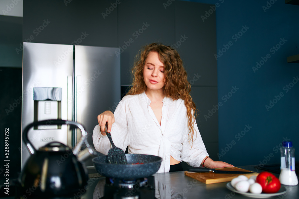 Young smiling curly caucasian woman cooks breakfast on a frying pan ...