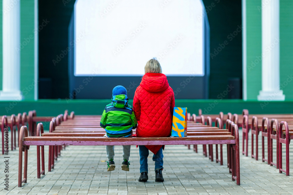Rear view of two viewers - a boy and his mother sitting on a bench ...