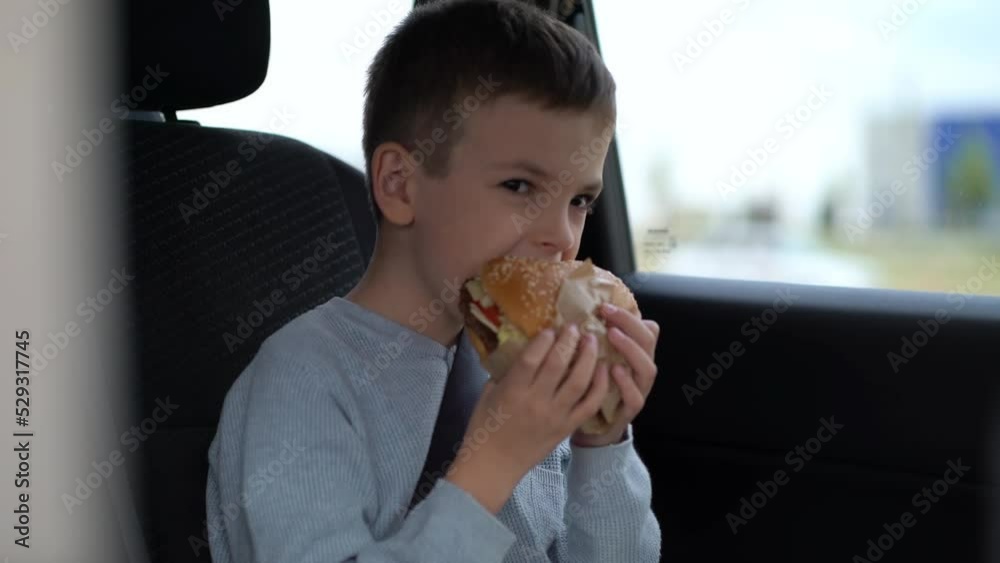 Boy child eating a big burger in the back seat of a car in the parking ...