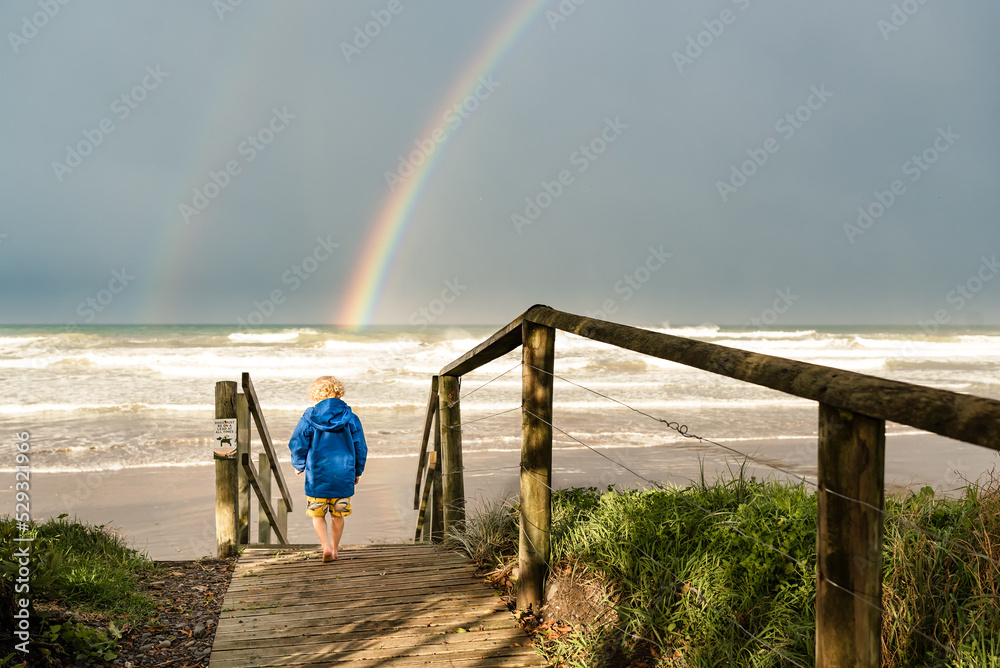 Young boy walking on boardwalk with a rainbow in the sky at a beach ...