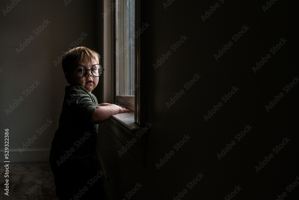 Toddler boy standing next to window in dark room wearing round glasses ...