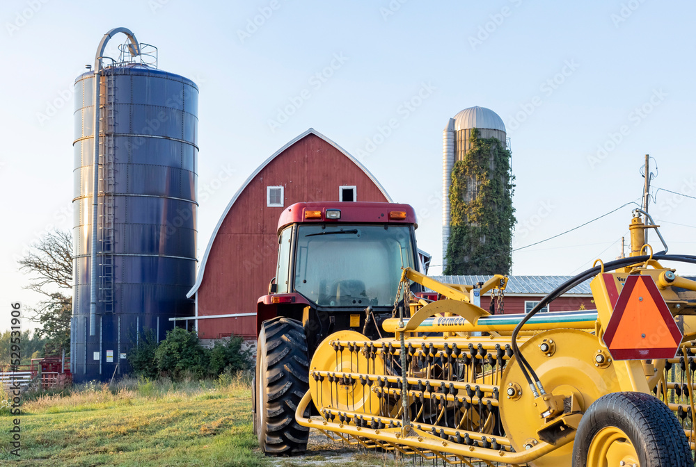 A Vermeer rake attached to a tractor in front of a red Gothic arch barn ...