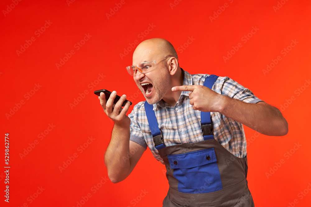 Angry bald man in work clothes and protective glasses shouts into the ...