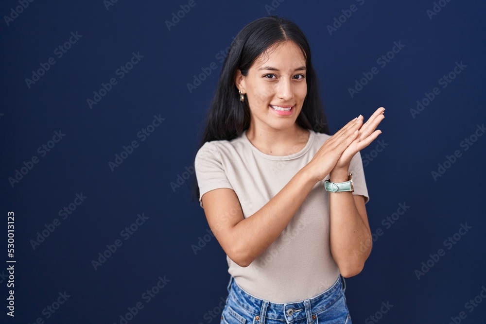 Young hispanic woman standing over blue background clapping and ...