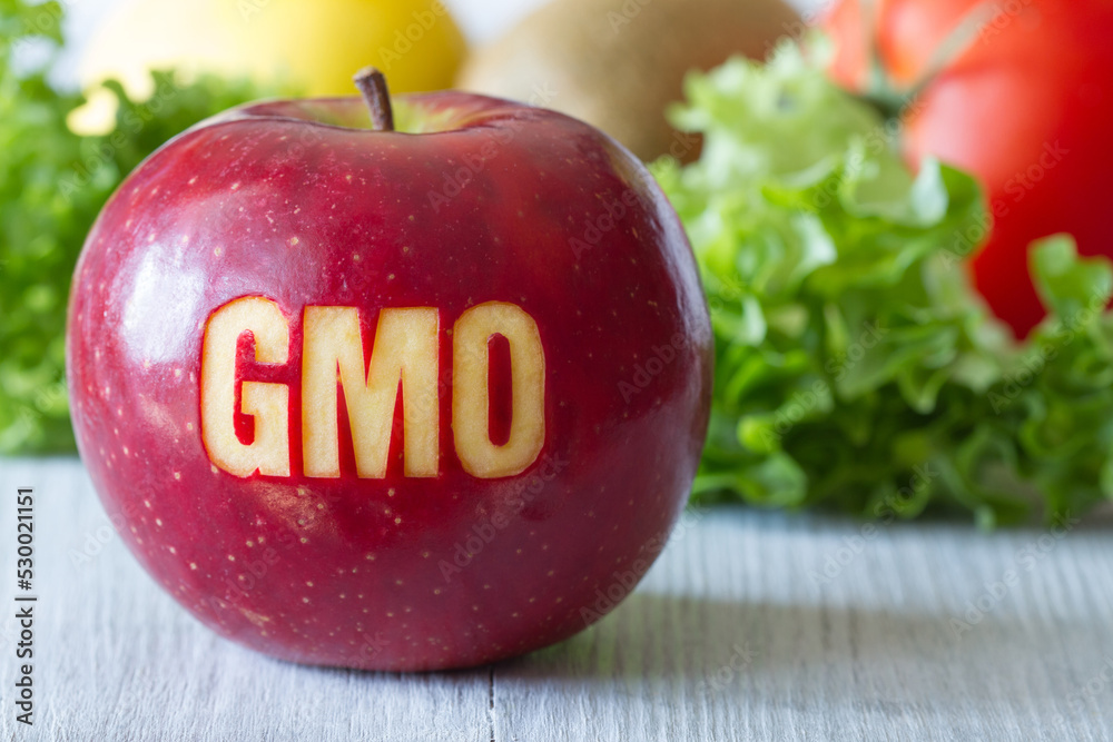 GMO text on red apple, vegetables and fruits in background, concept of ...