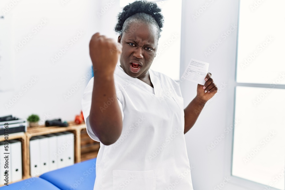Young african woman holding covid record card at clinic annoyed and ...