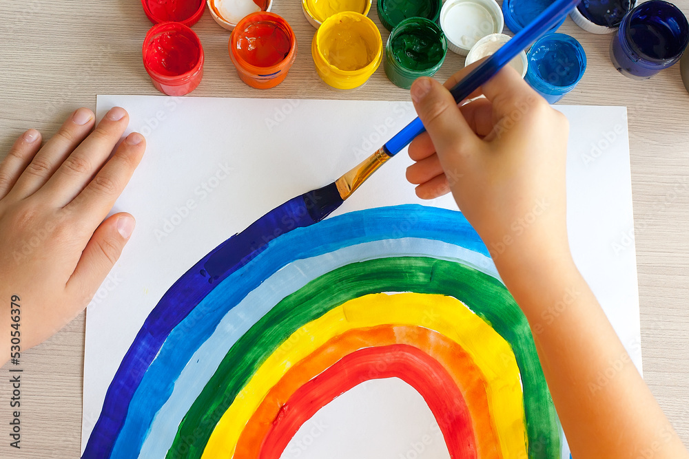 A child's hand draws a rainbow with a brush.Many cans of red yellow ...