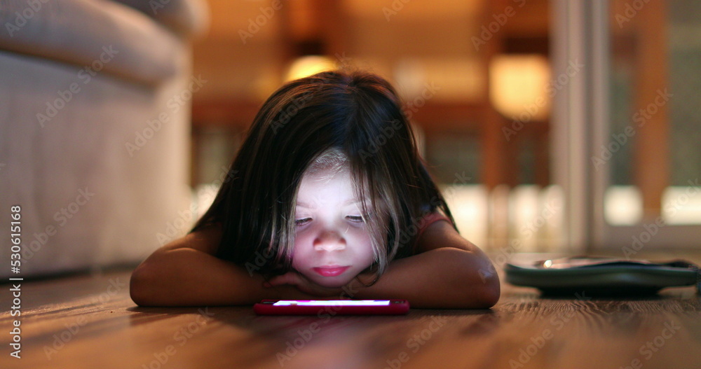 Little girl lying on floor at home looking at smartphone screen ...