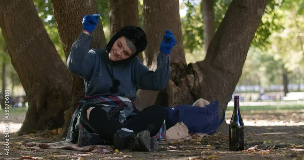 Portrait of drunken homeless woman sitting under tree in urban park ...