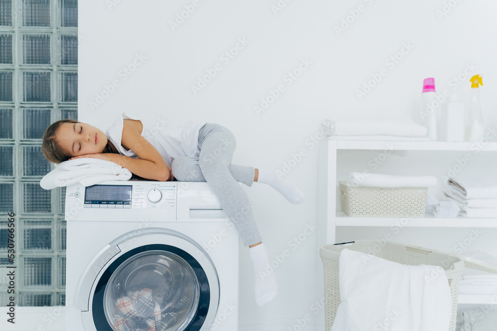 Horizontal shot of little girl has rest on washing machine, feels tired ...