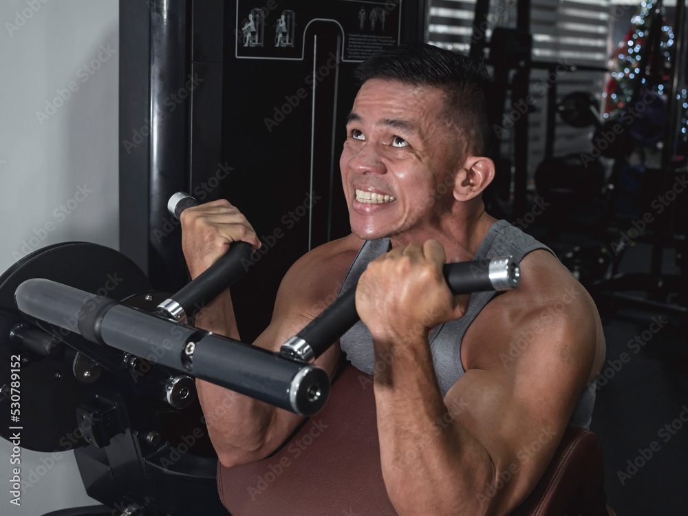 A brawny asian man grunts while doing some machine preacher curls at ...