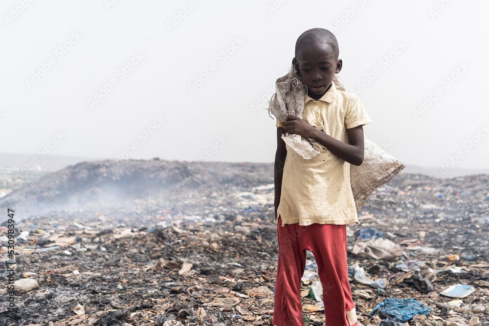 Neglected, malnourished African boy standing desolate in the midst of a smelly and smoking ...