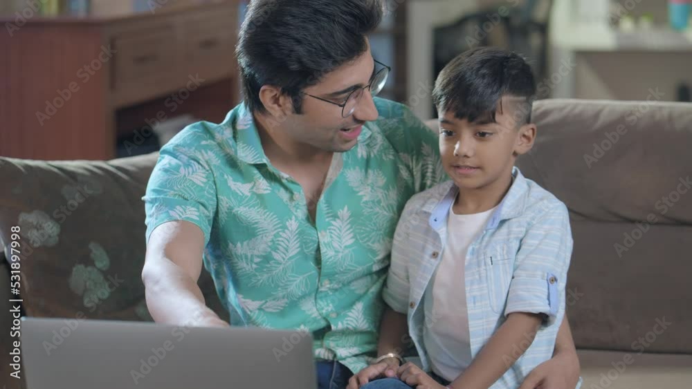 A young Indian Asian happy father and son duo is sitting on a couch ...