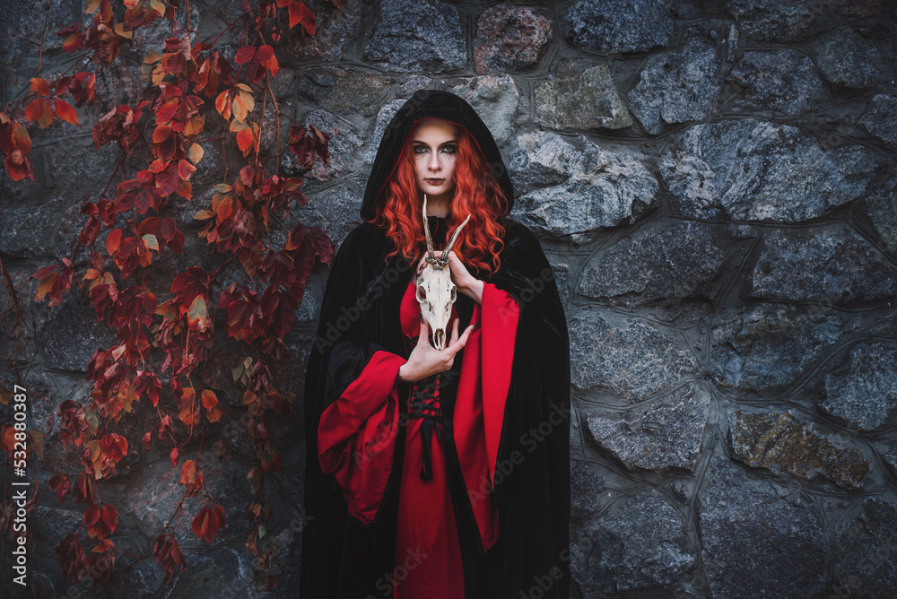 Red-haired beautiful young woman witch conjures and holds a goat skull ...