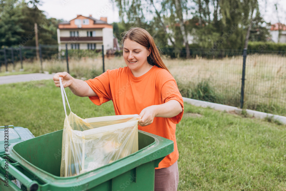 Young happy woman putting garbage in green trash bin on back yard ...