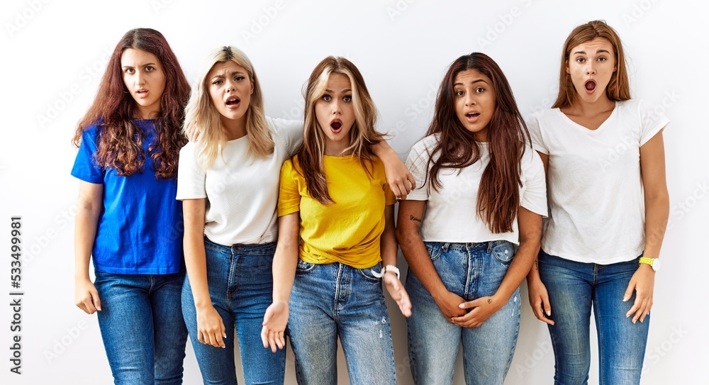 Group of young girl friends standing together over isolated background in shock face, looking ...