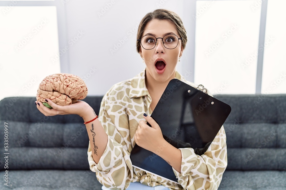 Young hispanic woman holding brain working at psychology clinic afraid ...