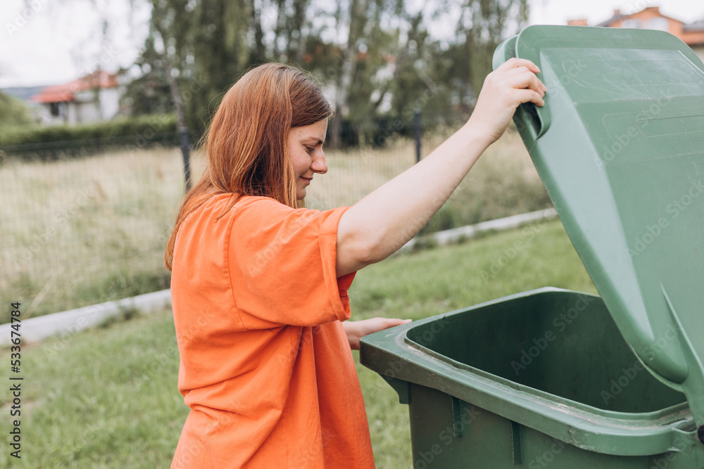 Young happy woman putting garbage in green trash bin on back yard ...