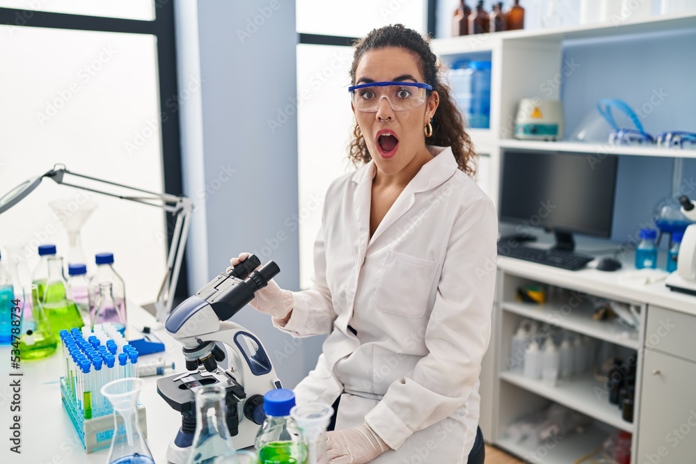 Young hispanic woman working at scientist laboratory scared and amazed ...