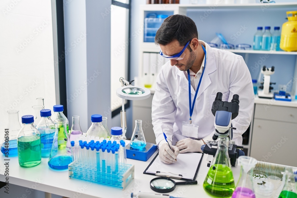 Young hispanic man scientist measuring liquid writing report at ...