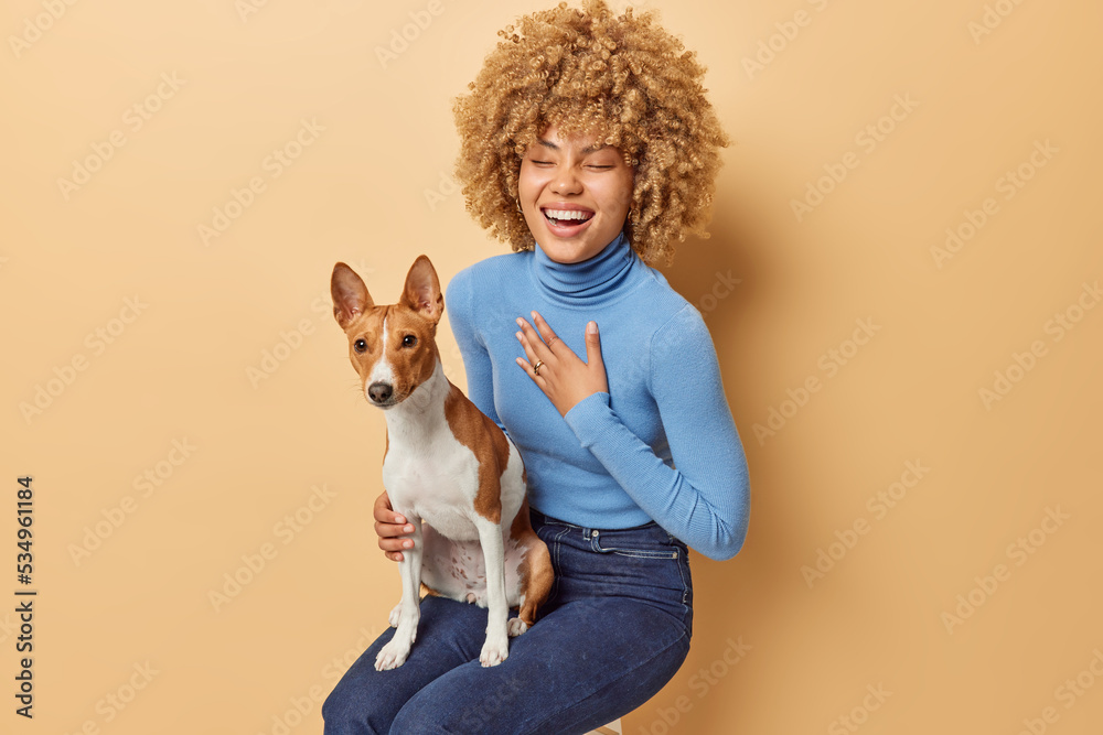Horizontal shot of overjoyed curly haired pet owner keeps hand on chest ...