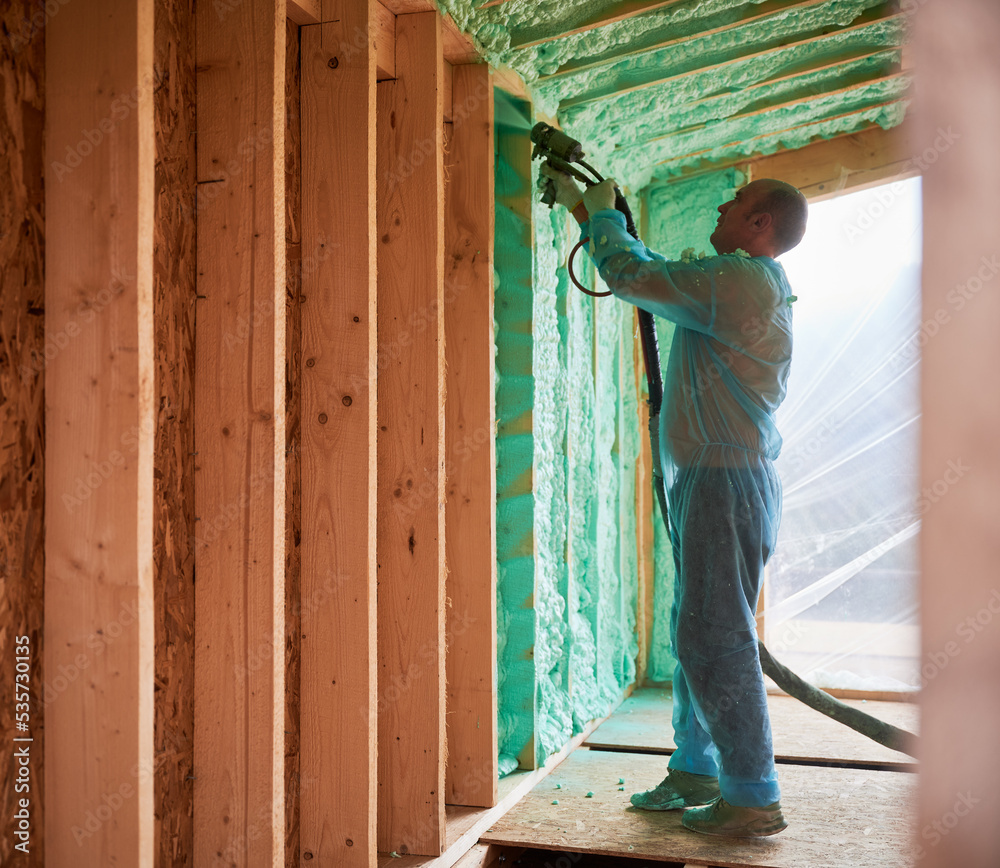 Male builder insulating wooden frame house. Man worker spraying ...