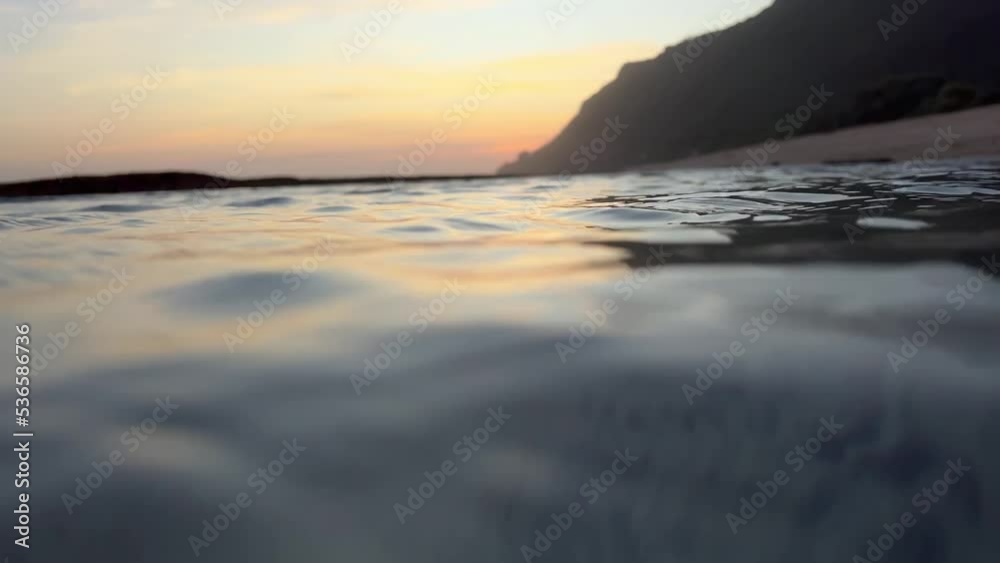Sunset on a wild beach underwater. Ocean ripples and dark reef rocks on ...