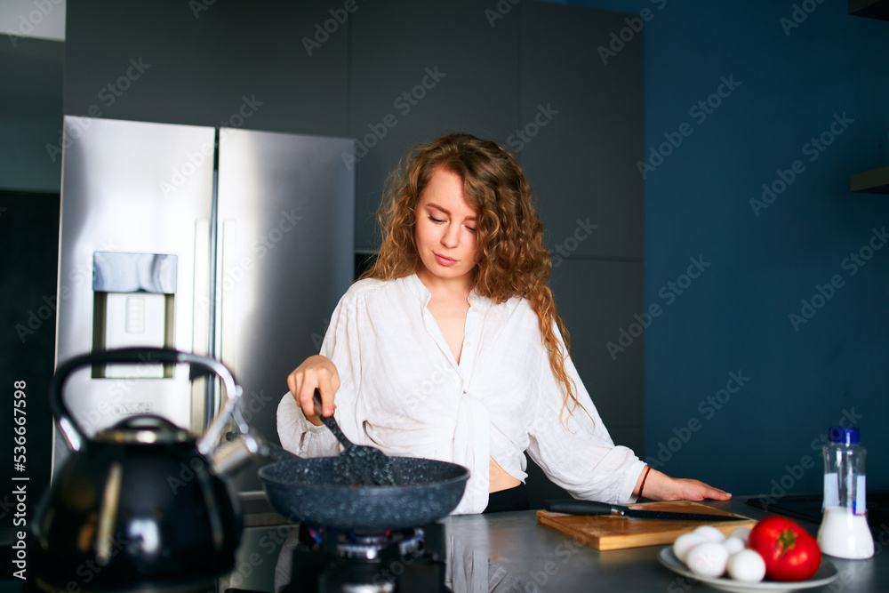 Young smiling curly caucasian woman cooks breakfast on a frying pan ...