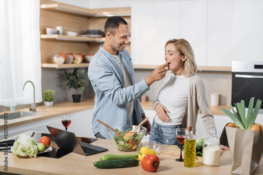 Cheerful married multinational couple using laptop while cooking ...