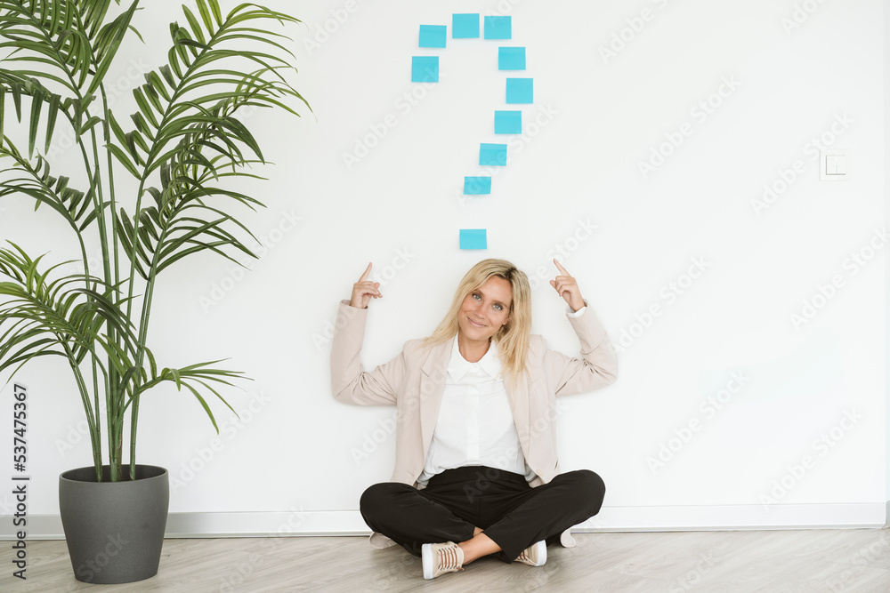 Businesswoman sitting on the floor in office pointing at question mark ...