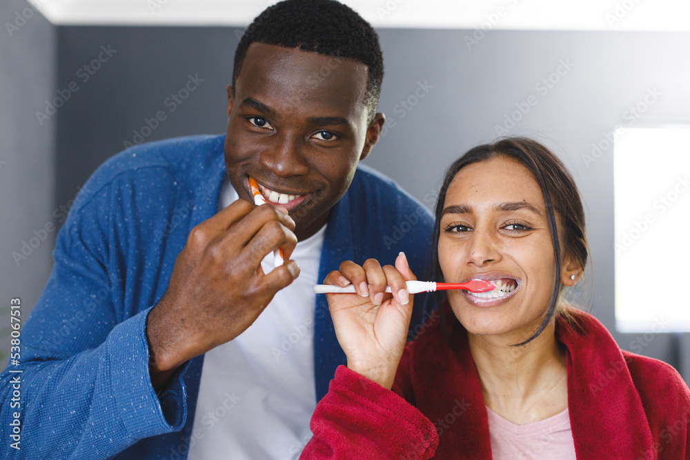 Happy diverse couple in brushing teeth in bathroom, looking to camera ...