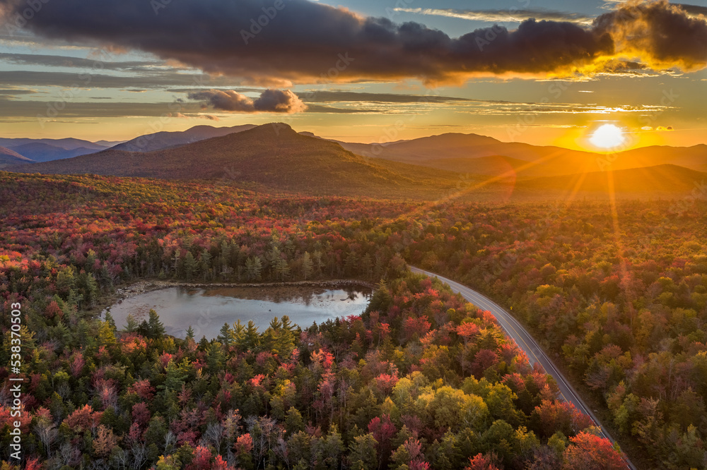 Sunrise over a scene of a road and lake through a forest with trees in ...