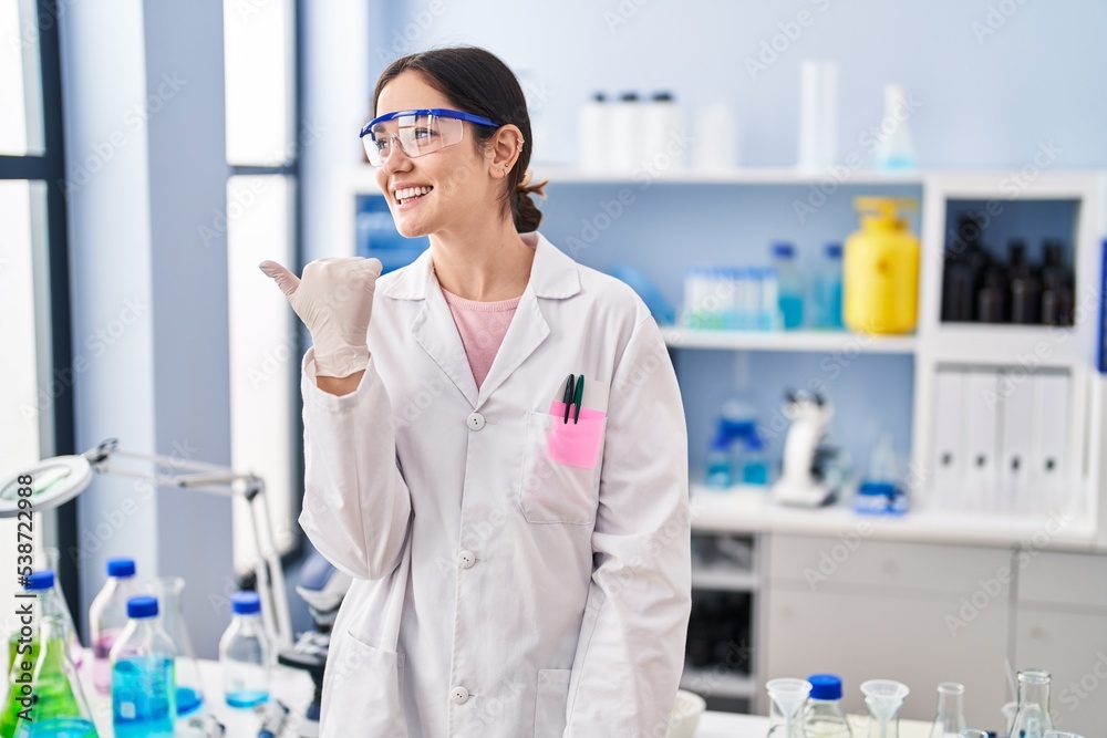 Young brunette woman working at scientist laboratory pointing thumb up ...
