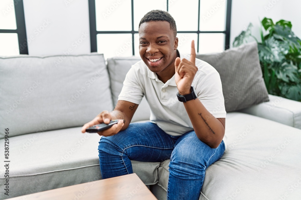 Young african man watching tv holding television remote control smiling ...