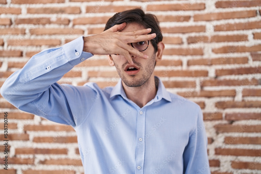Young hispanic man standing over brick wall background peeking in shock ...