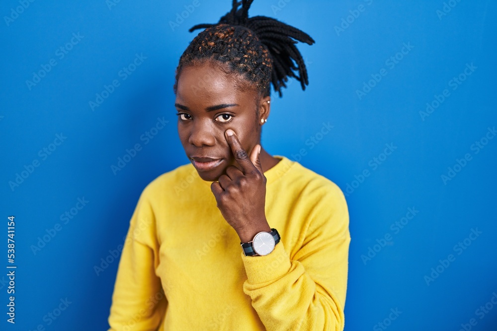Beautiful black woman standing over blue background pointing to the eye ...
