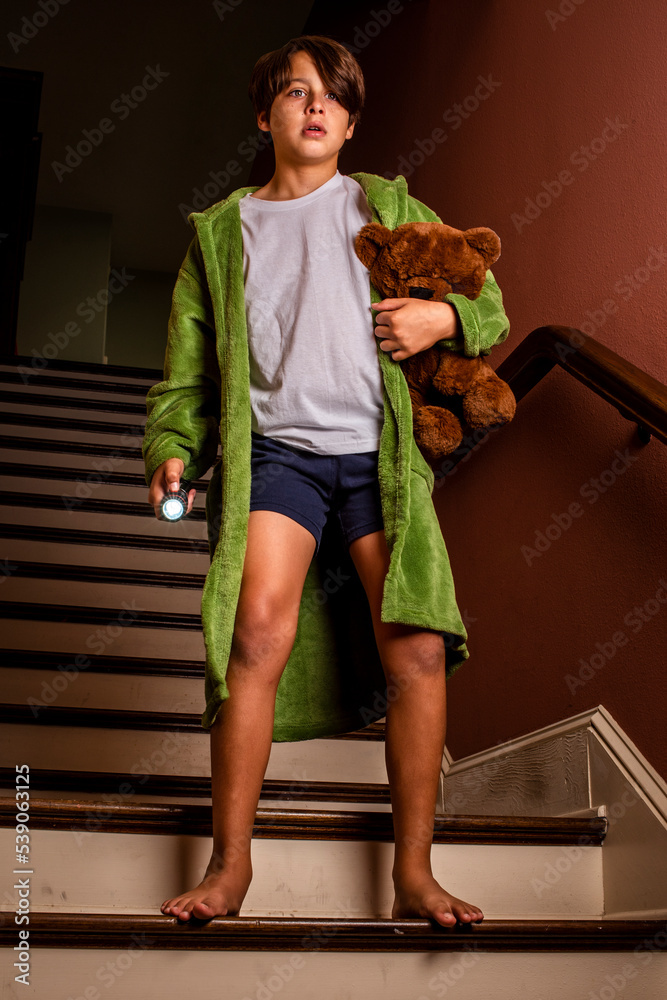 Scared young boy clutching his teddy bear and a flashlight on stairs at ...