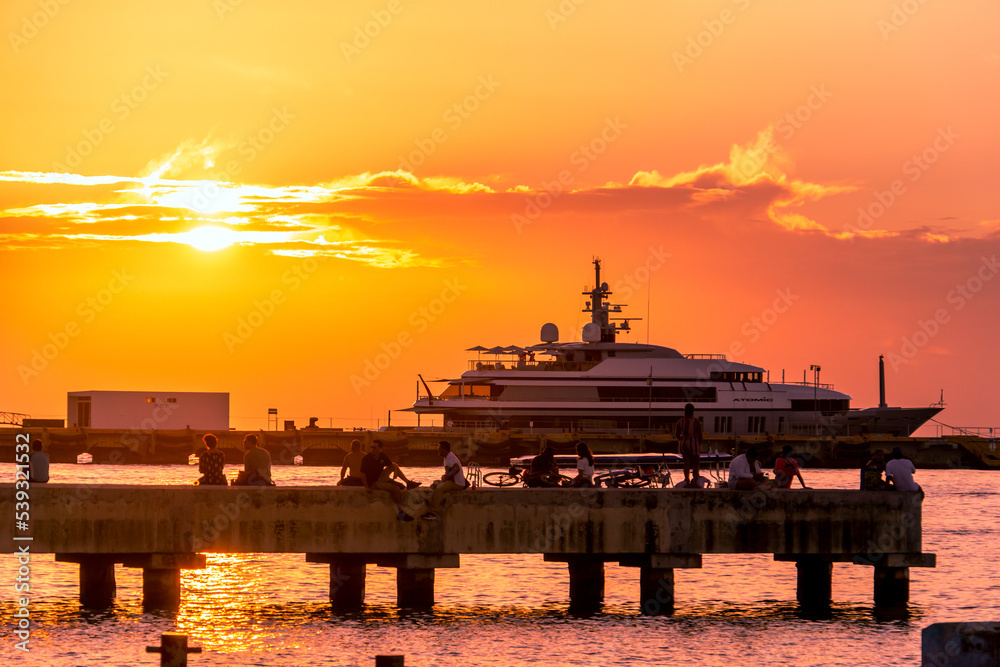 Port at sunset. Figures of people sit on the pier and look at the ...