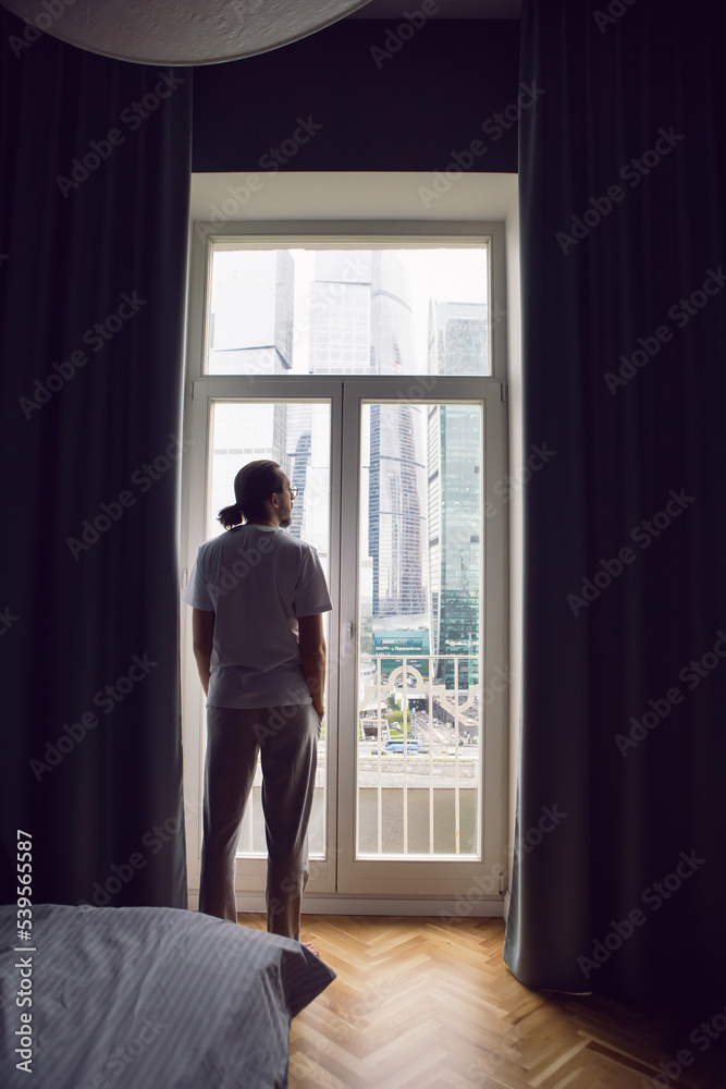 man with long hair, beard and glasses stands at a large window in an ...