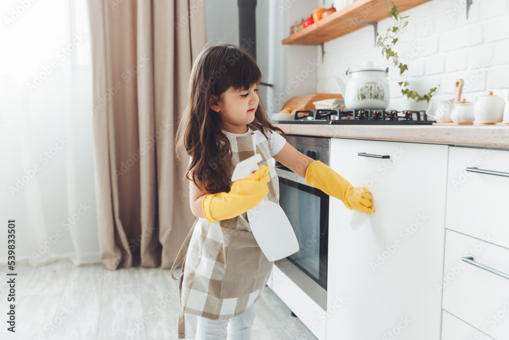 A cute little girl in an apron wipes the cupboard in the kitchen ...