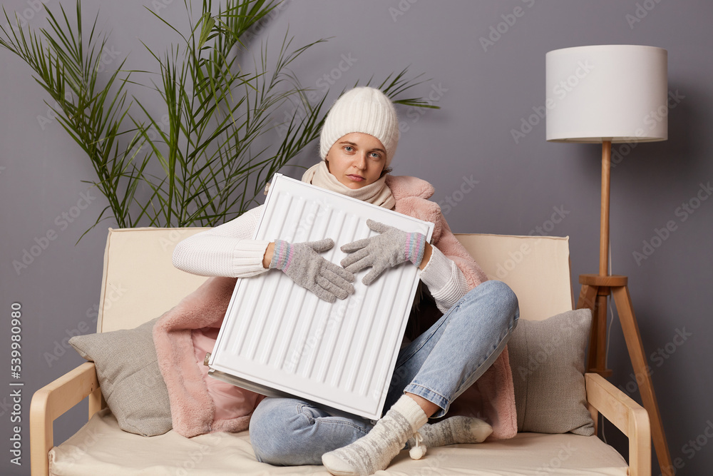 Indoor shot of sad young adult woman in coat and hat sit in cold living ...
