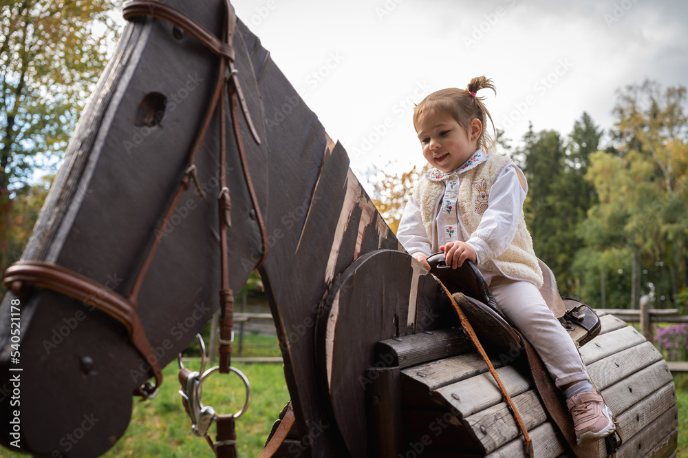 Little child girl riding on a wooden toy horse in a green park, autumn ...