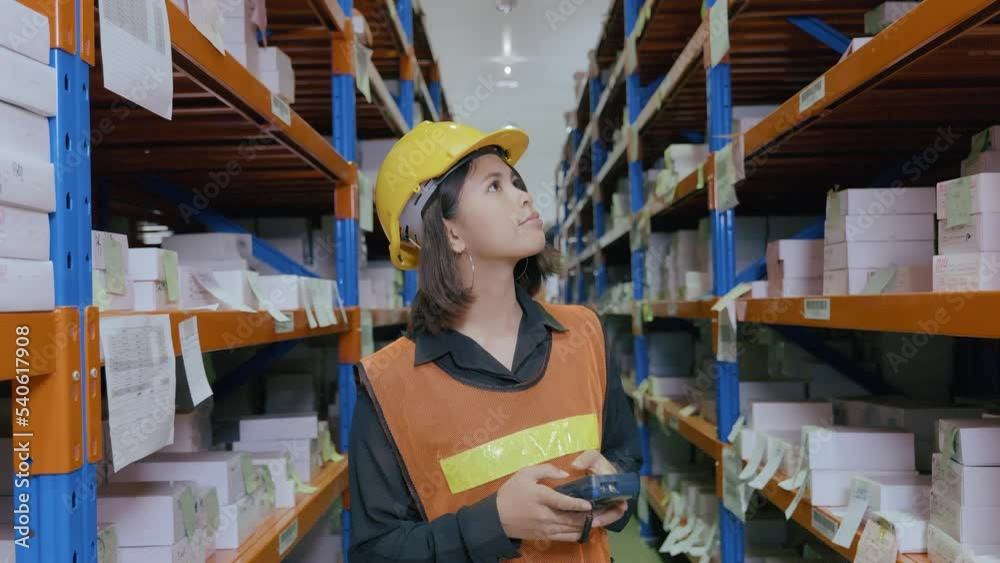 A female worker in uniform with her safety helmet on is walking by holding scanner for scanning ...