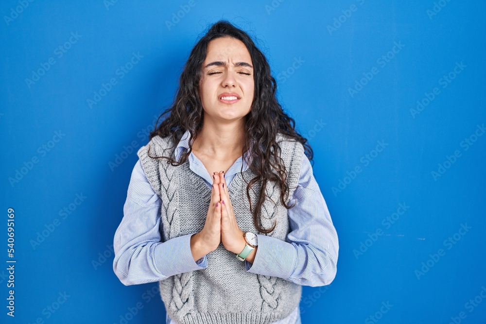 Young brunette woman standing over blue background begging and praying ...