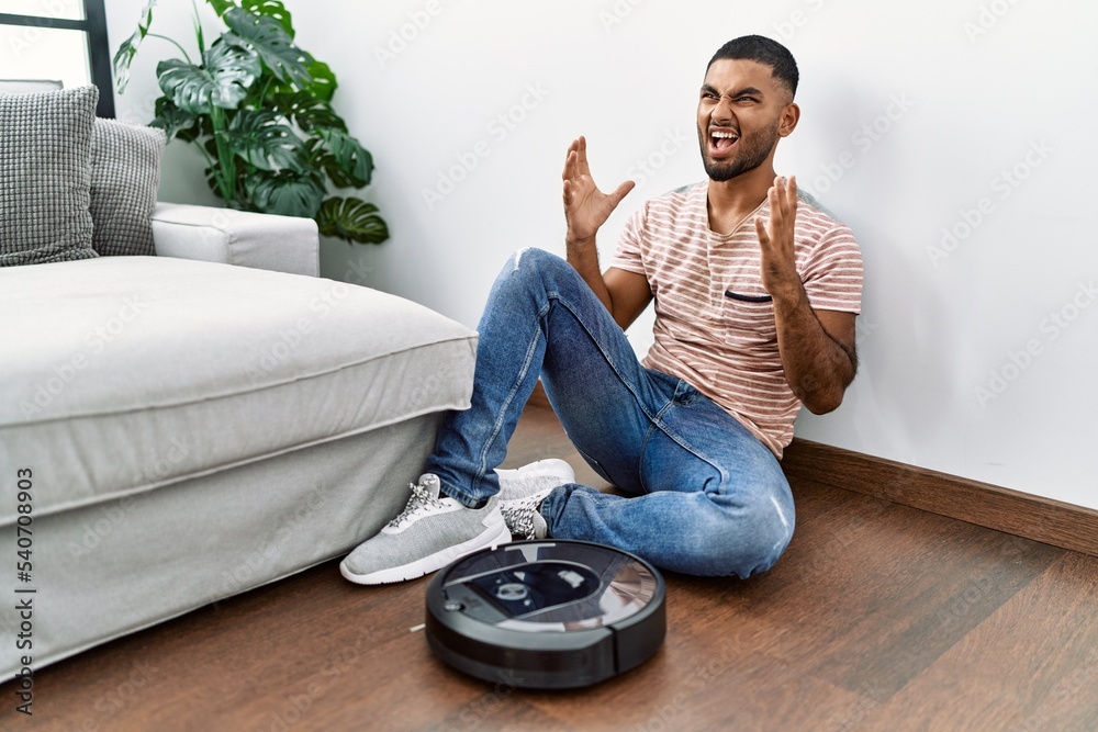 Young indian man sitting at home by vacuum robot crazy and mad shouting ...