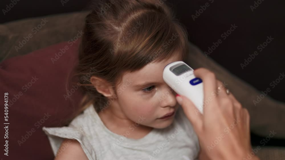 Caring mother measures her baby girl's temperature using a no-touch infrared kid thermometer ...