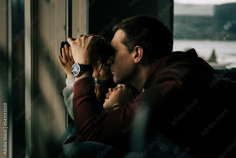 father and daughter looking out the window with binoculars at nature ...